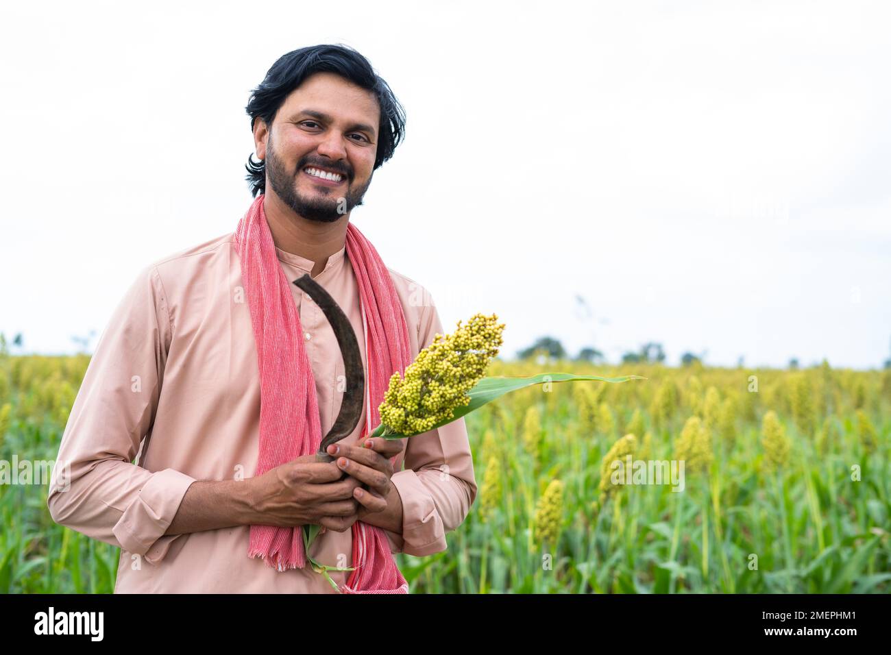 Happy smiling young farmer holding crop and sickle by looking camera at corn or maize field ...