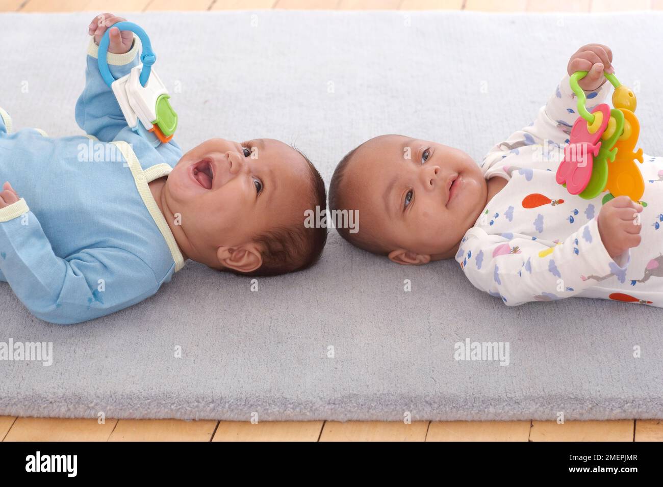 Twin boys (4 months) lying on a mat on the floor, both holding plastic ...
