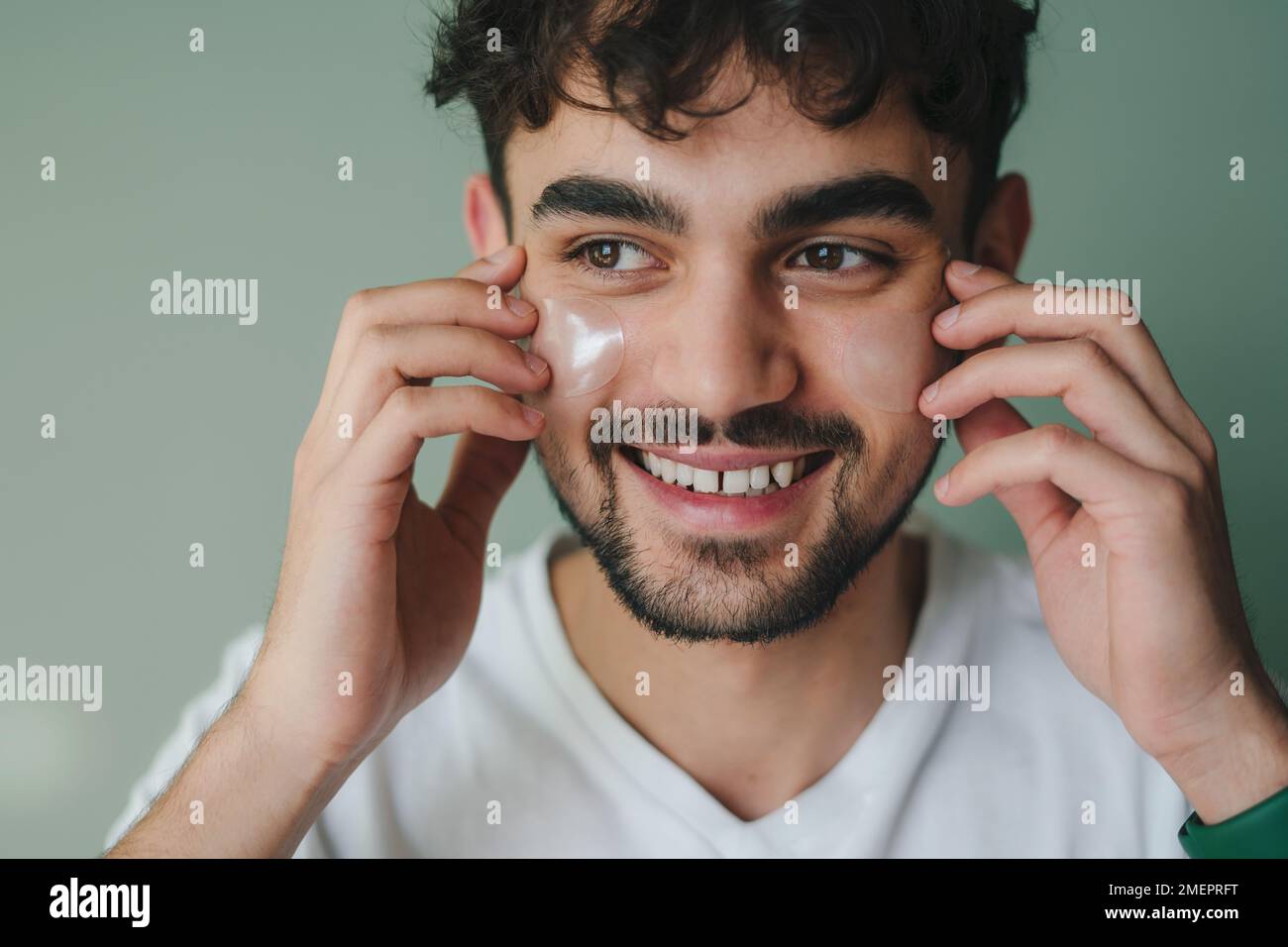 Portrait of smiling man looking away applying moisturizer cream on his face isolated over green ...