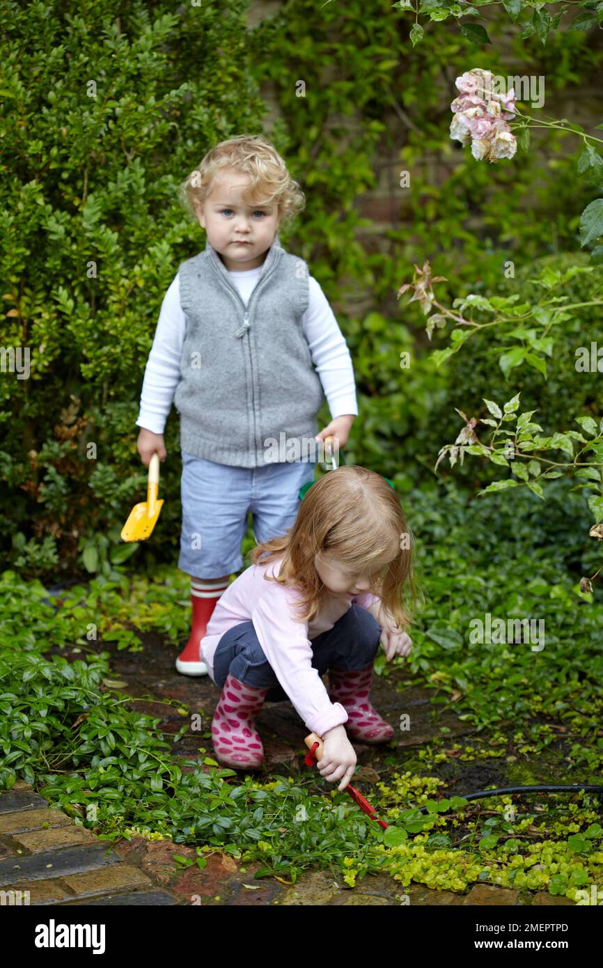 Boy holding bucket and spade standing behind young girl crouched on ...