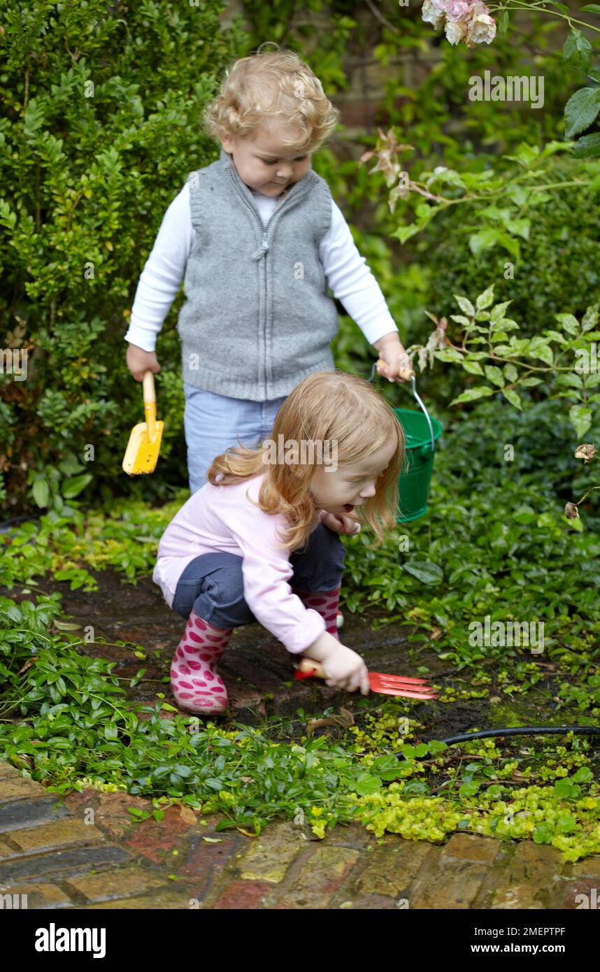 Boy holding bucket and spade standing behind young girl crouched on ...