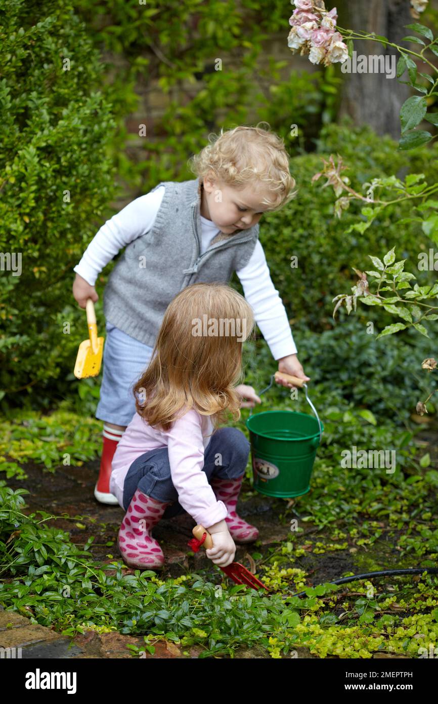 Boy holding bucket and spade standing behind young girl crouched on ...