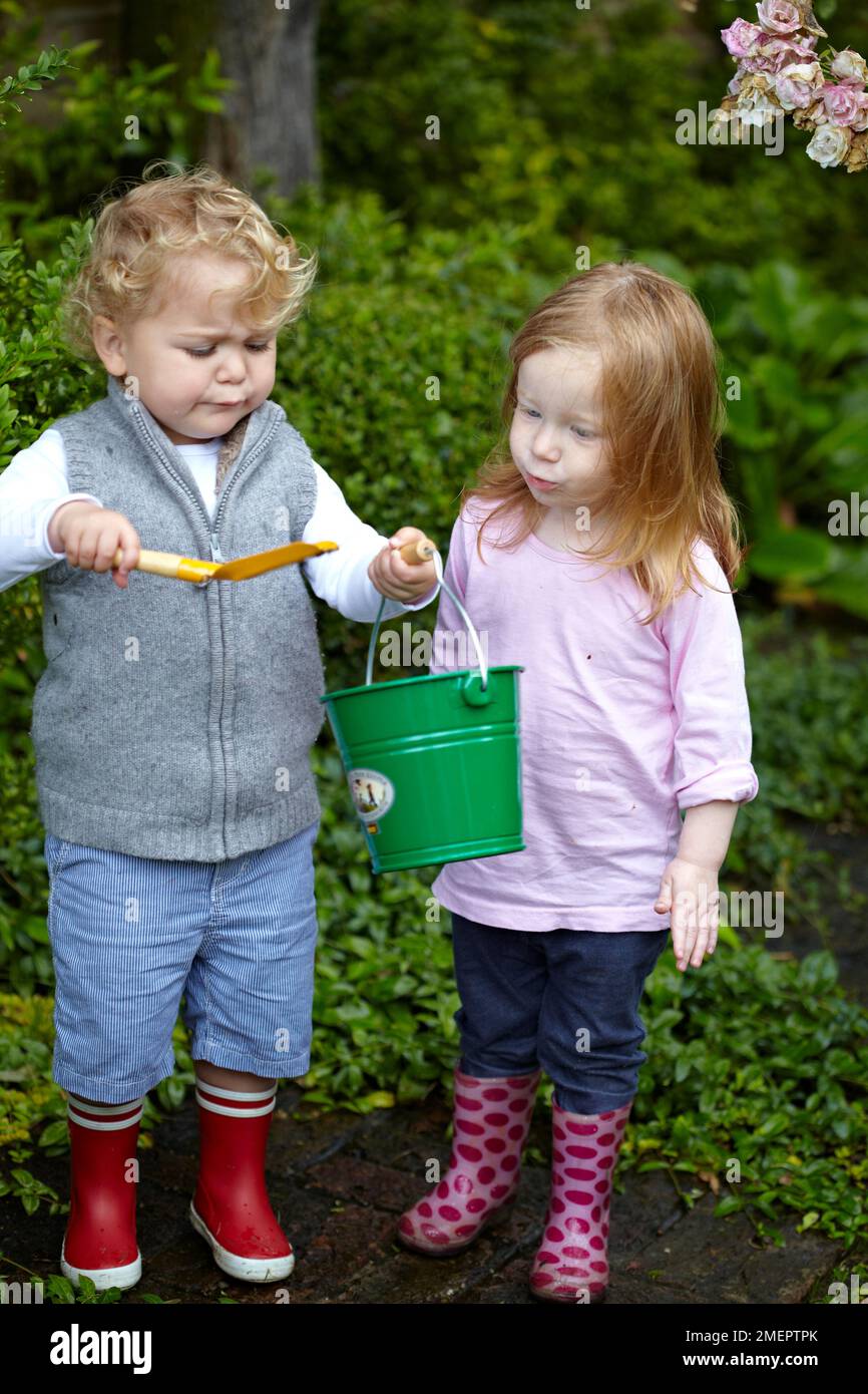 Boy holding bucket and spade standing next to young girl, 2 years 4 ...