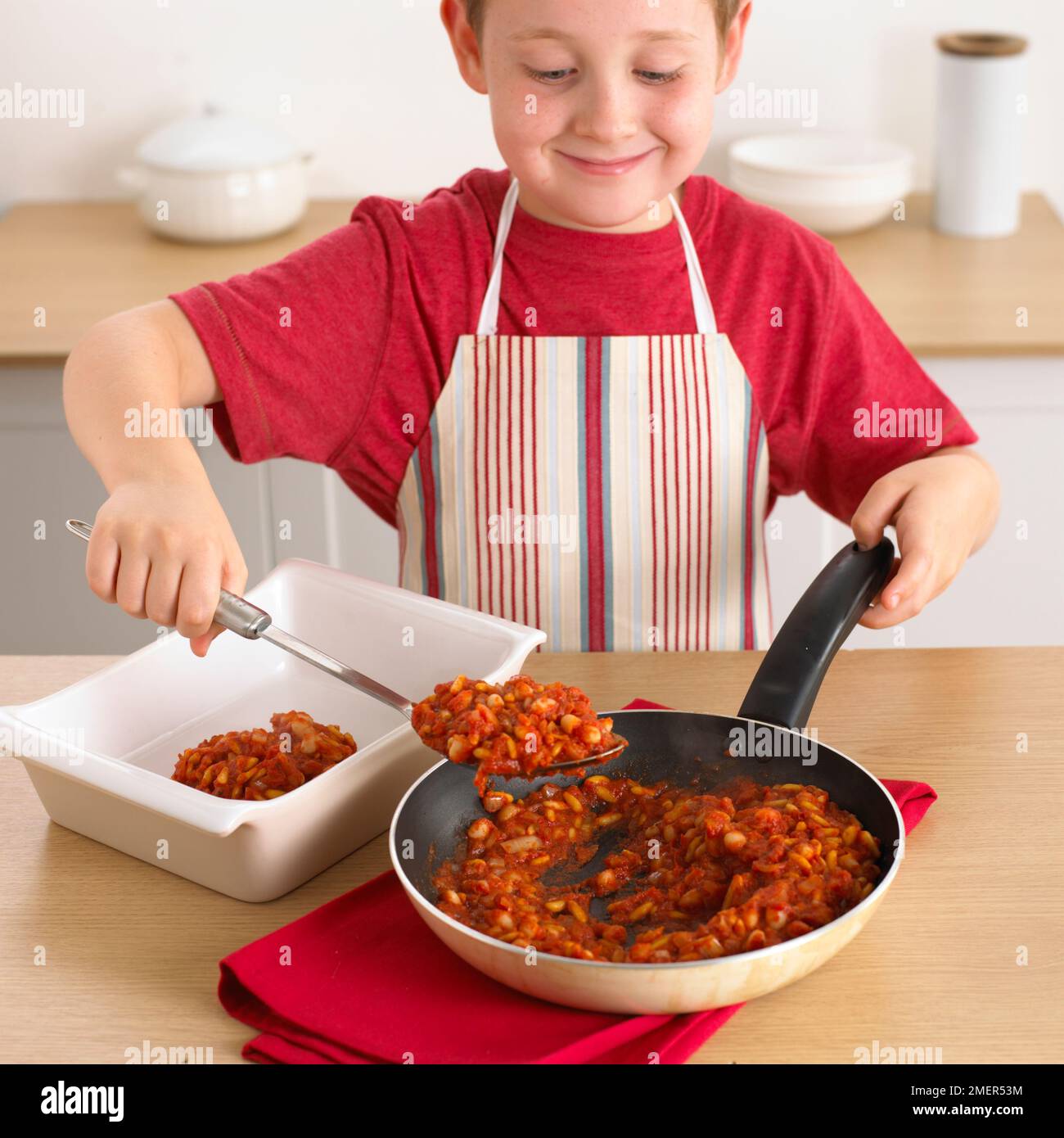 Boy placing bean mixture from frying pan into oven dish, 8 years Stock ...