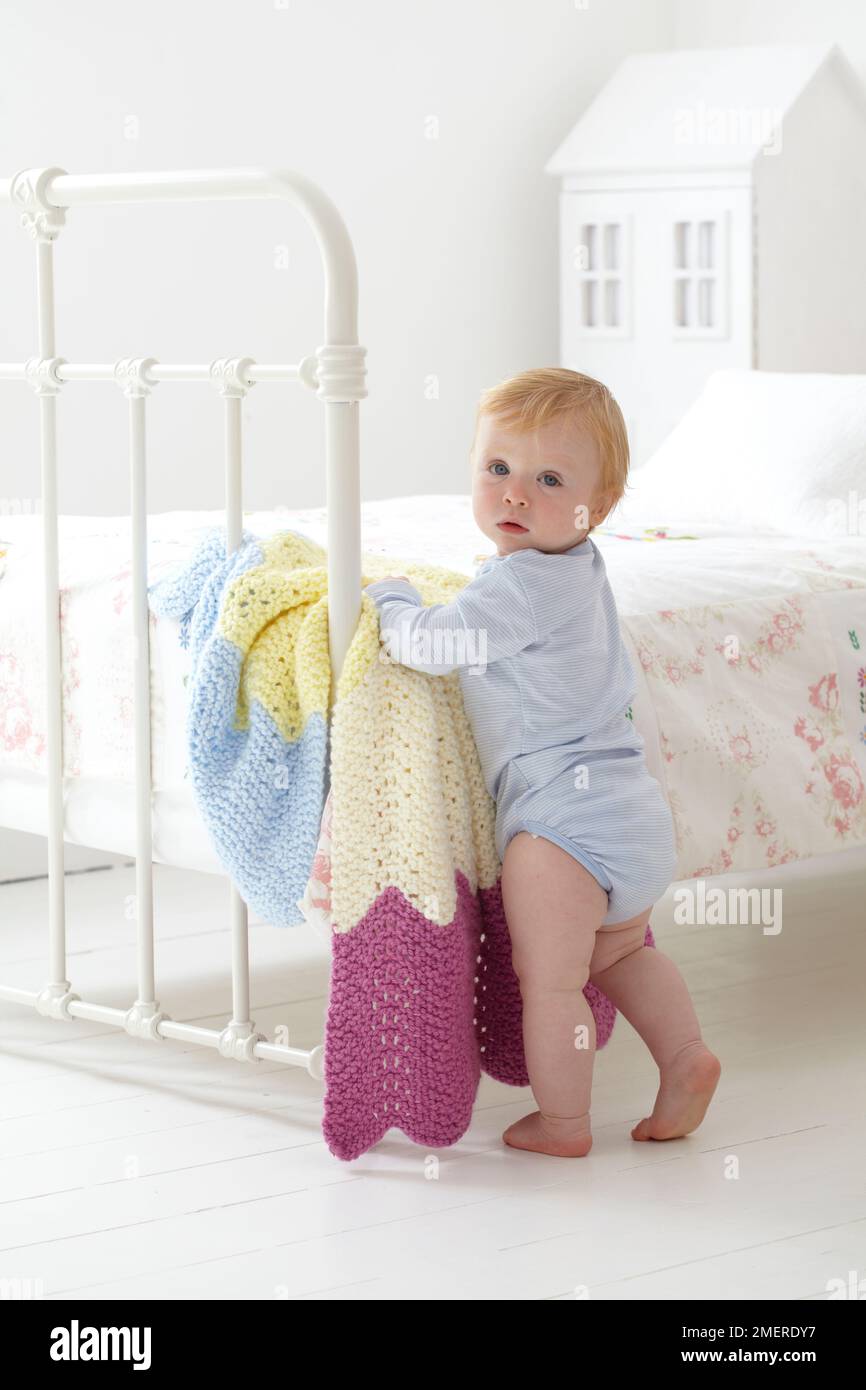 Baby boy standing next to bed with knitted blanket draped over the edge ...