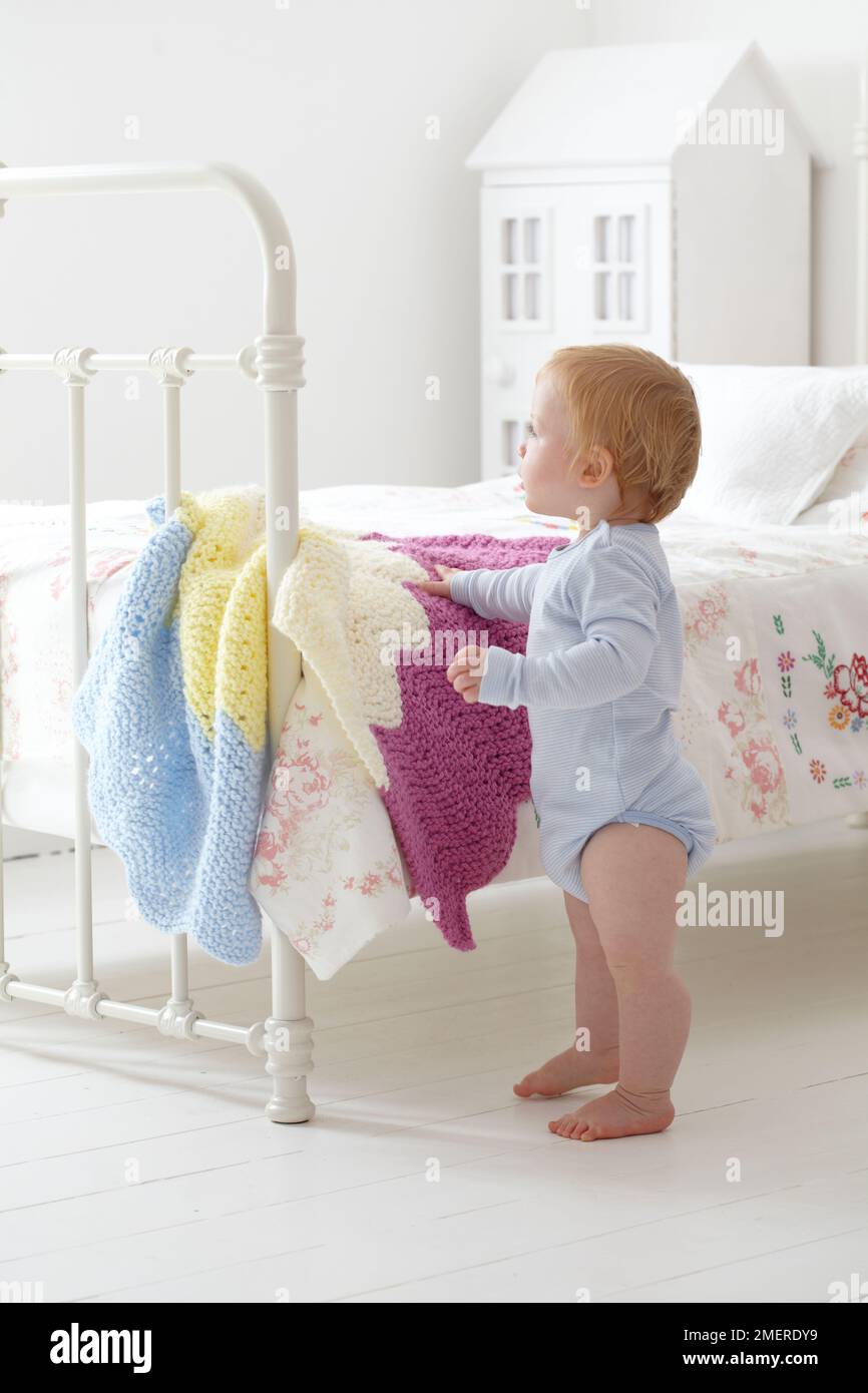Baby boy standing next to bed with knitted blanket draped over the edge ...