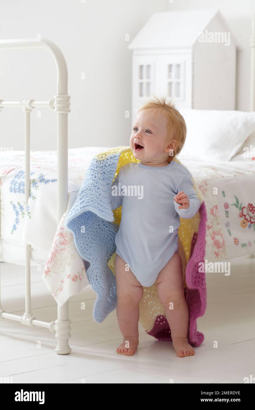 Baby boy standing next to bed holding blanket over shoulders, 12 months ...