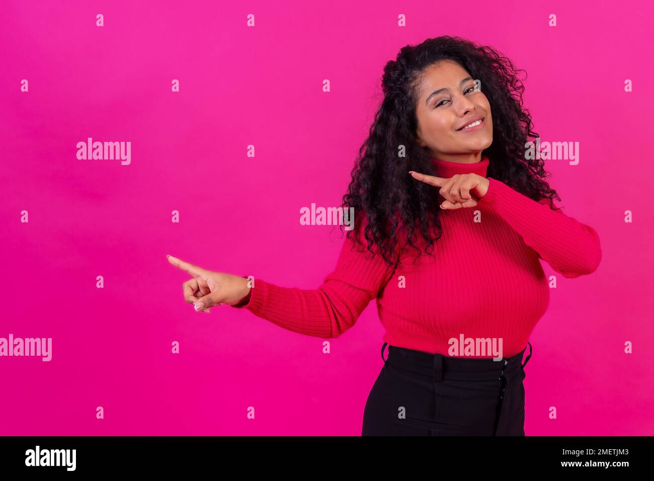 Curly-haired woman pointing left on a pink background, studio shot ...