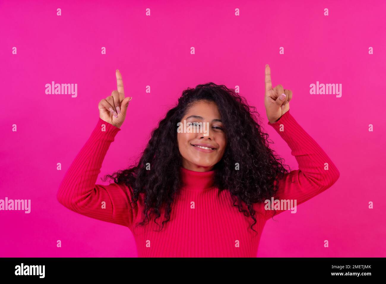 Curly-haired woman pointing up on a pink background, studio shot, copy ...