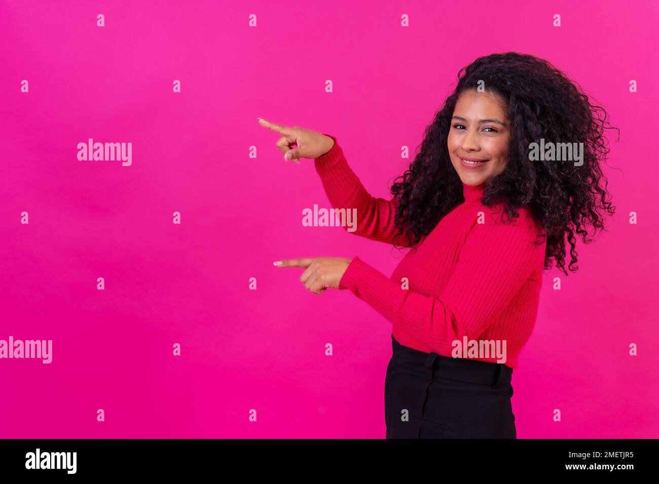 Curly-haired woman pointing left on a pink background, studio shot ...