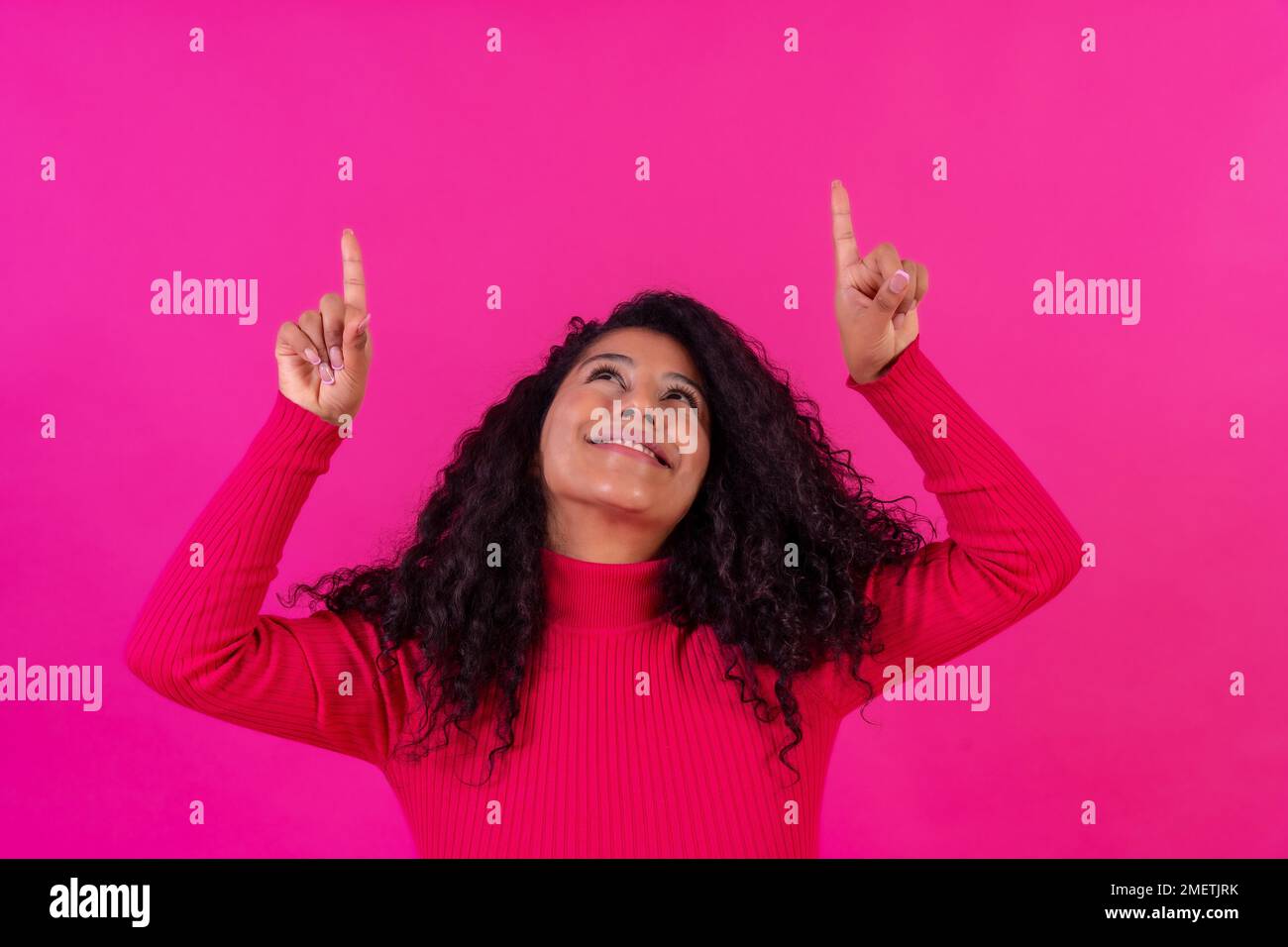 Curly-haired woman pointing up on a pink background, studio shot, copy ...