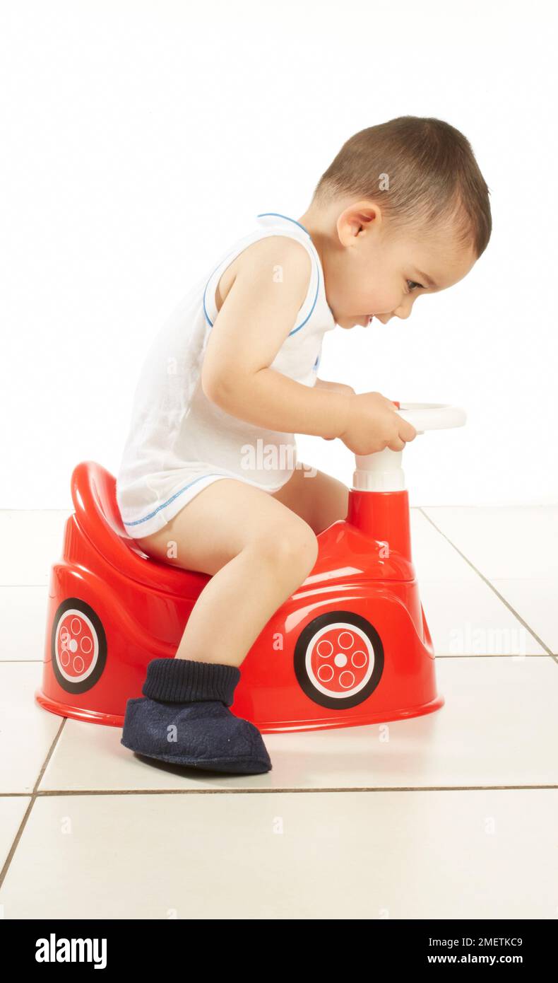 Boy wearing vest and slippers sitting on red car potty with steering wheel, 15 months Stock ...