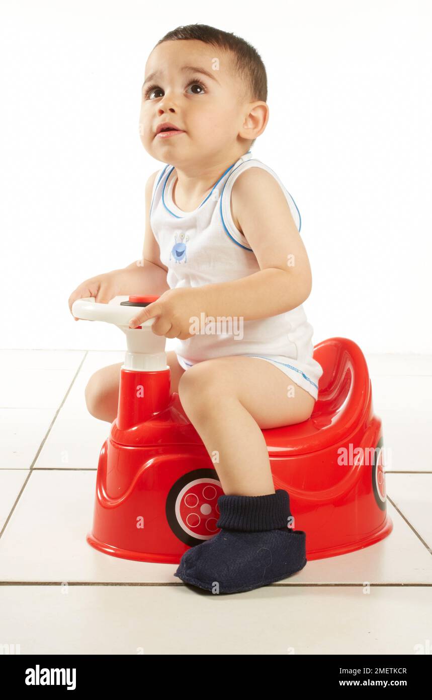 Boy wearing vest and slippers sitting on red car potty with steering wheel, 15 months Stock ...