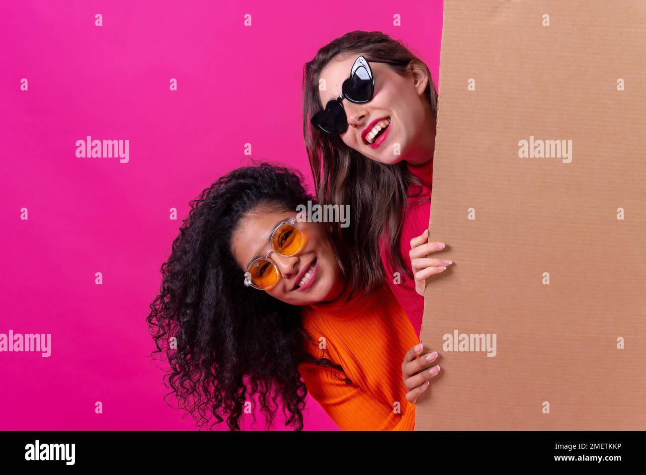 Two smiling beautiful girl friends hold a cardboard sign on a pink ...