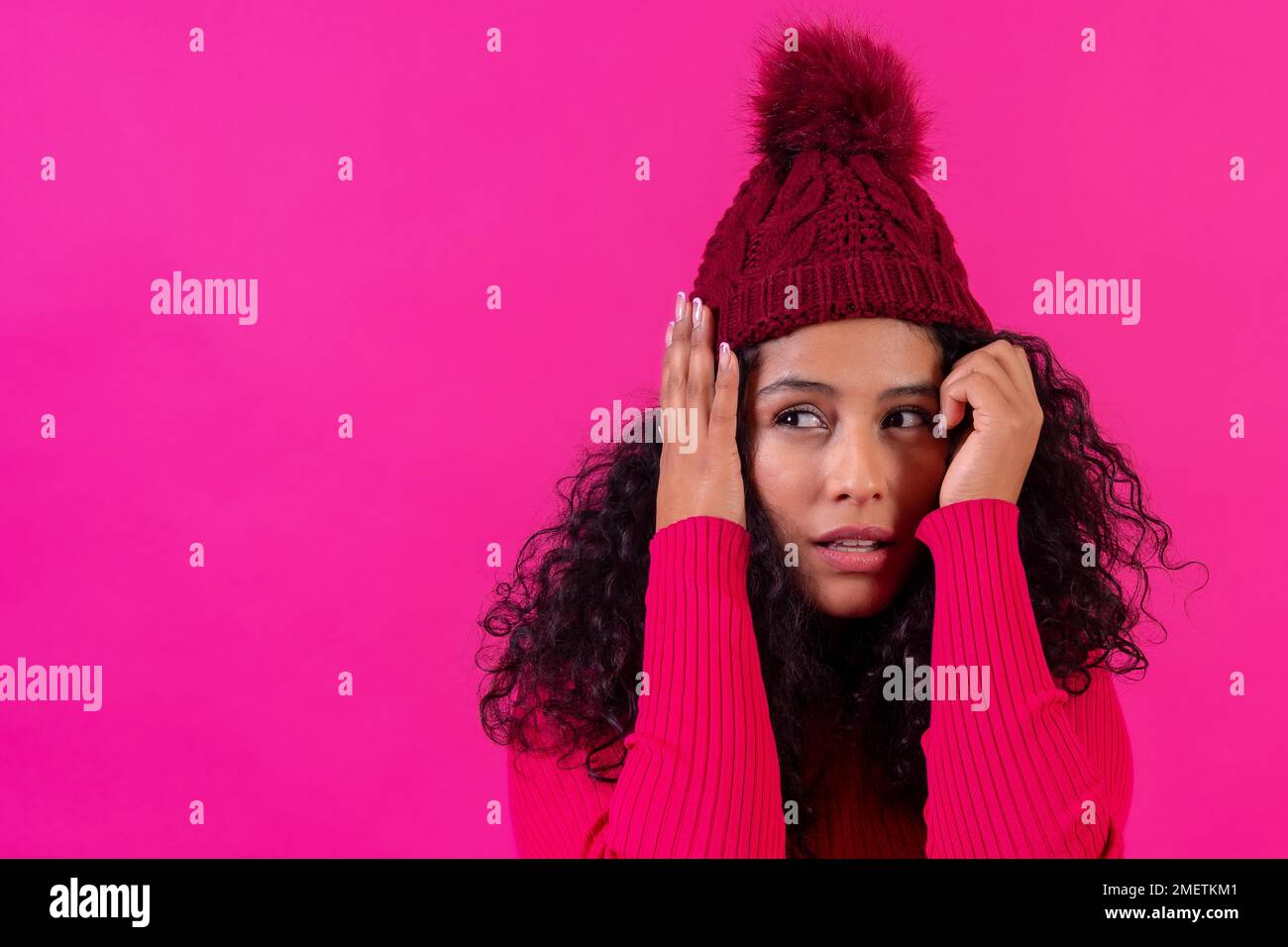 Curly-haired woman in a wool cap on a pink background scared, studio ...