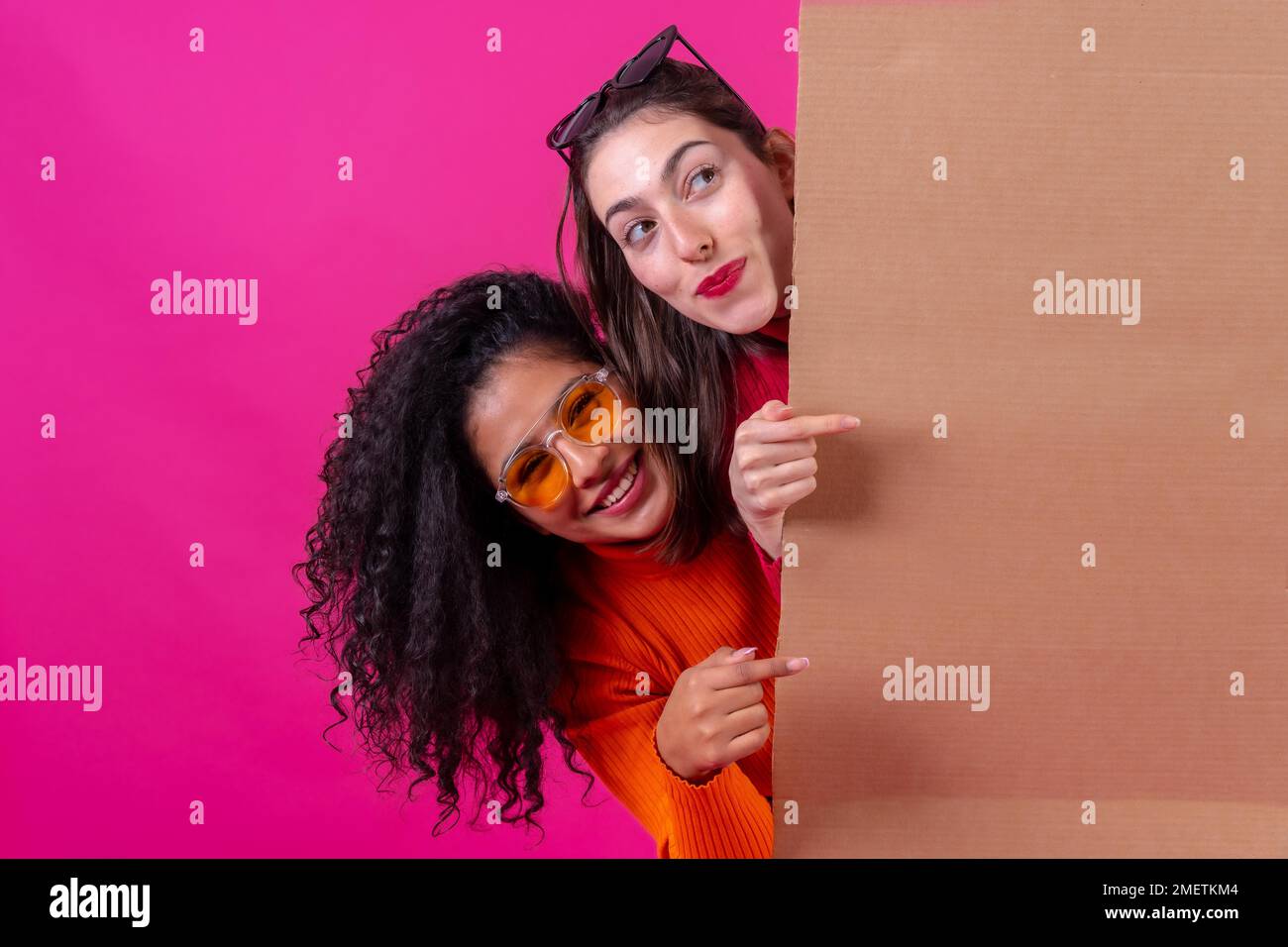 Two smiling beautiful female friends pointing at cardboard sign over ...