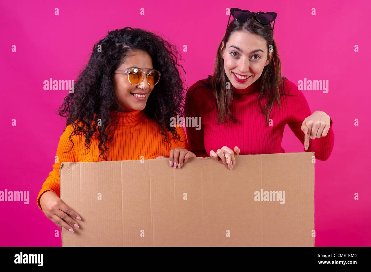 Two multiethnic female friends smiling and pointing at cardboard sign ...
