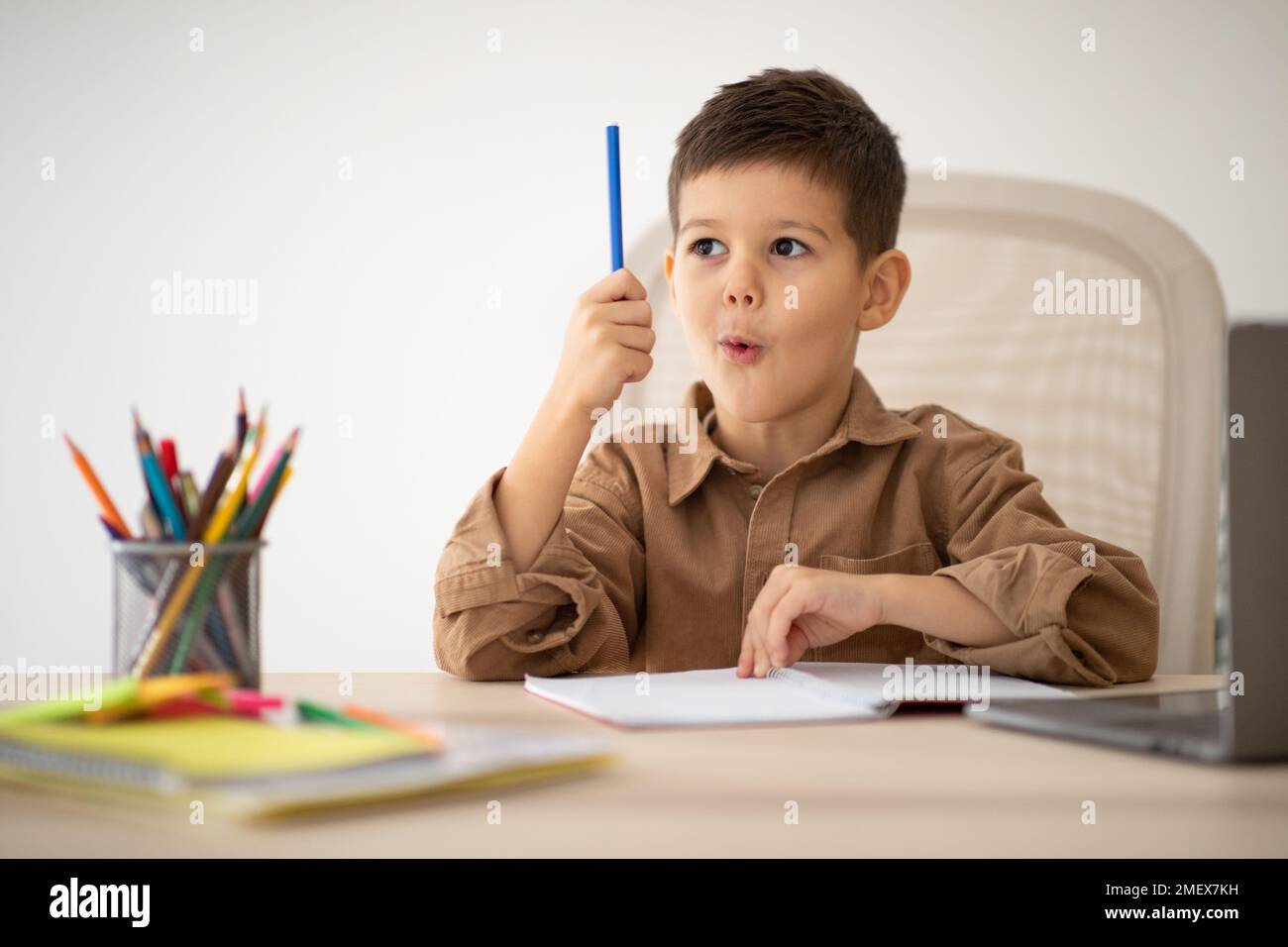 Cheerful cute little boy sits at table, studies, looks at pencil and ...