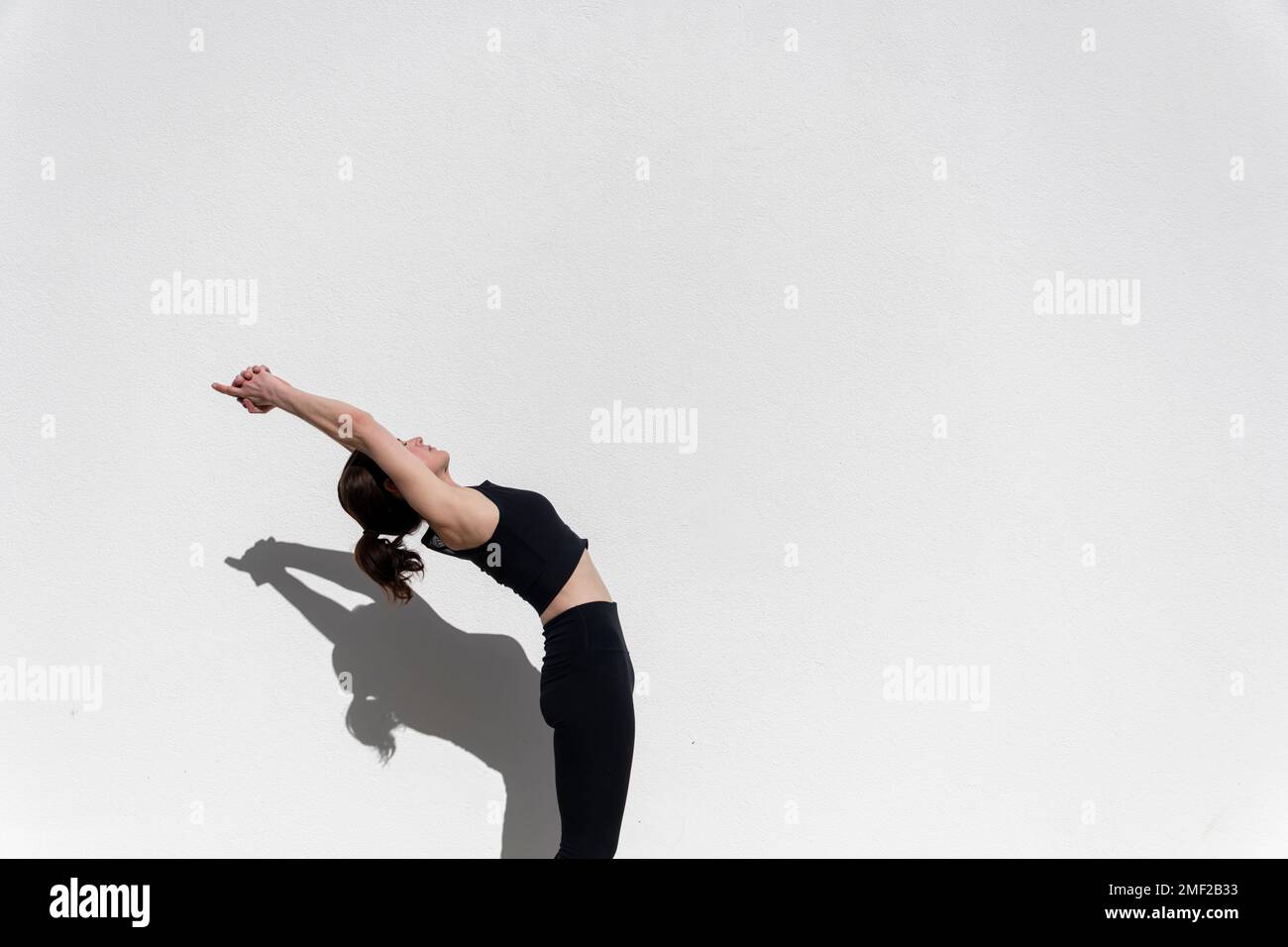 Woman doing a mountain pose with backbend, yoga outdoors, white wall ...