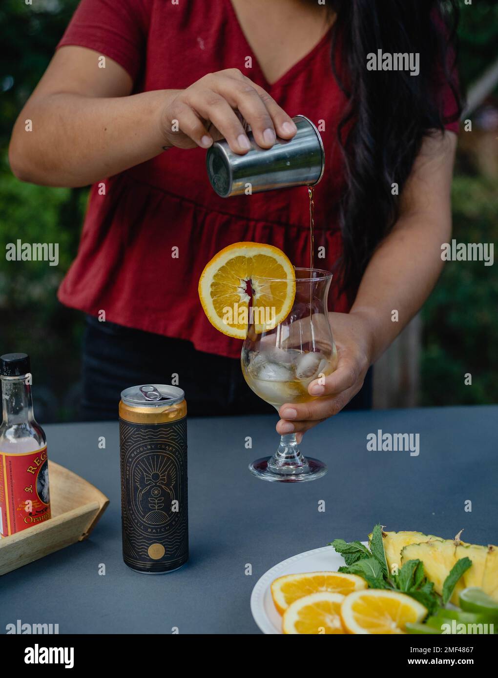 A bartender who is a woman of color pours alcohol into a cocktail glass with an orange slice ...