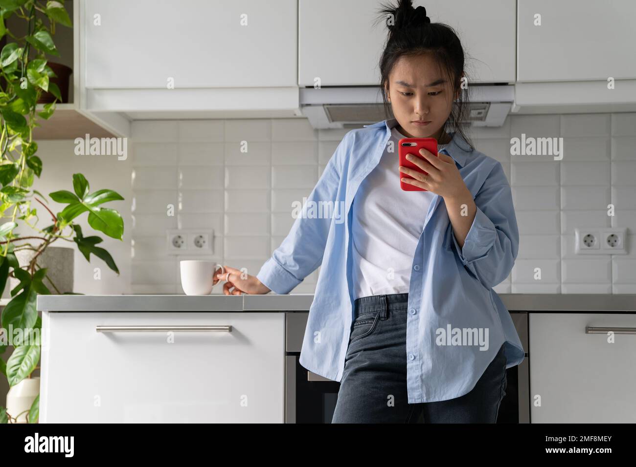 Displeased embarrassed Asian woman with phone standing in kitchen reads ...