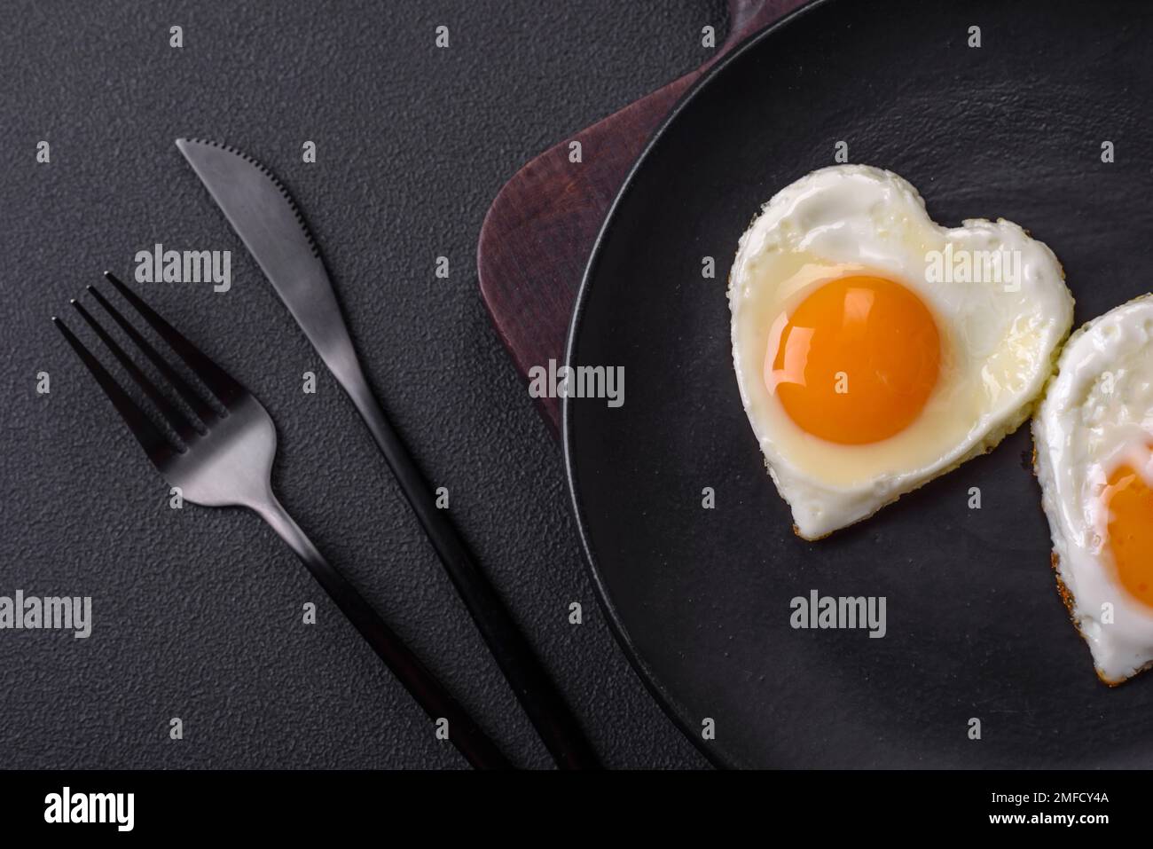 Two heart-shaped fried eggs on a black ceramic plate on a dark concrete ...