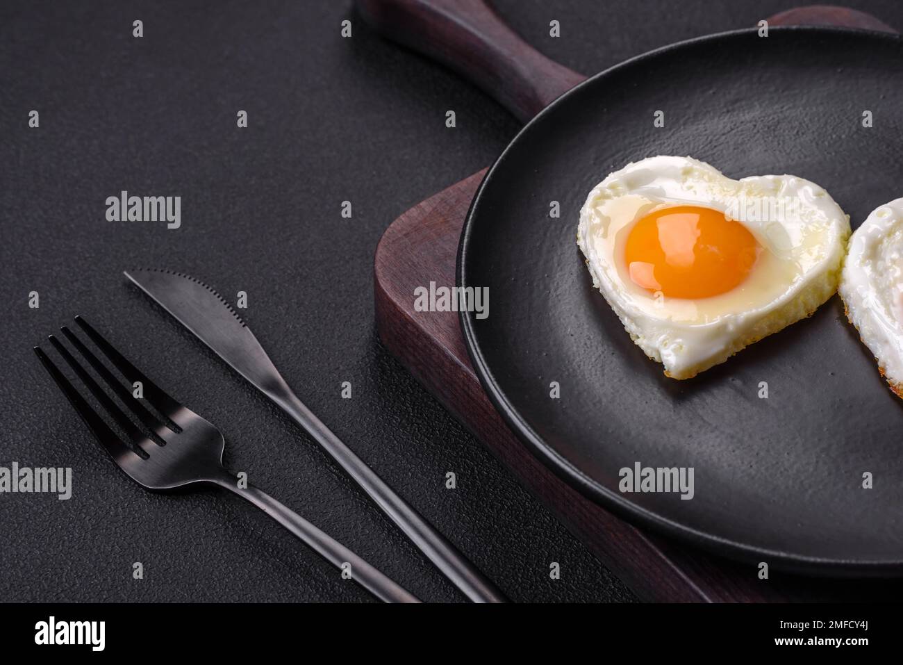 Two heart-shaped fried eggs on a black ceramic plate on a dark concrete ...
