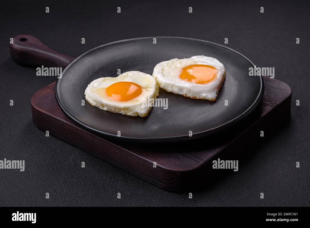 Two heart-shaped fried eggs on a black ceramic plate on a dark concrete ...
