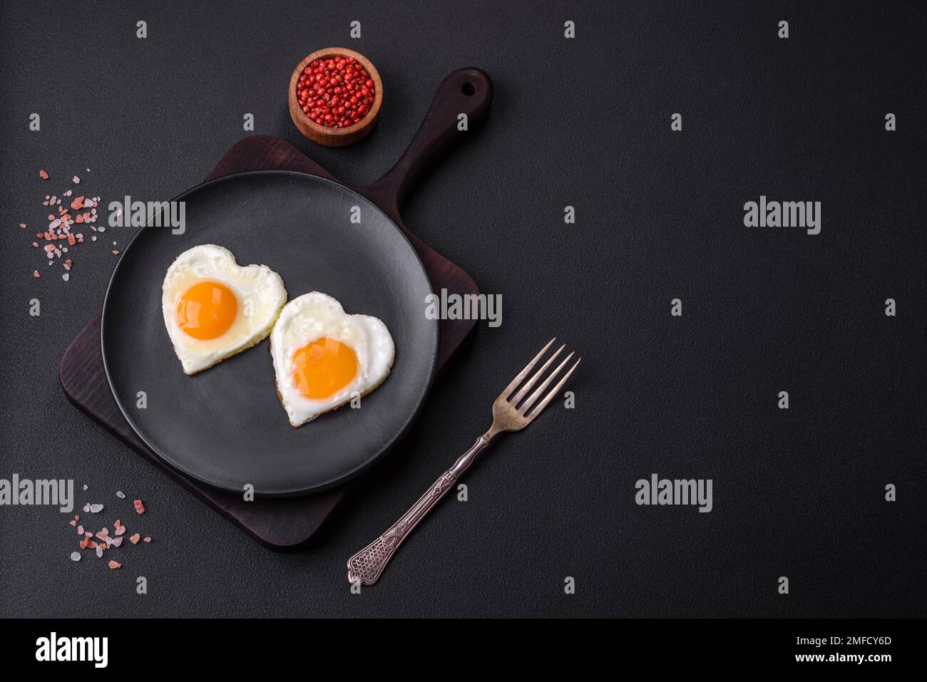 Two heart-shaped fried eggs on a black ceramic plate on a dark concrete ...