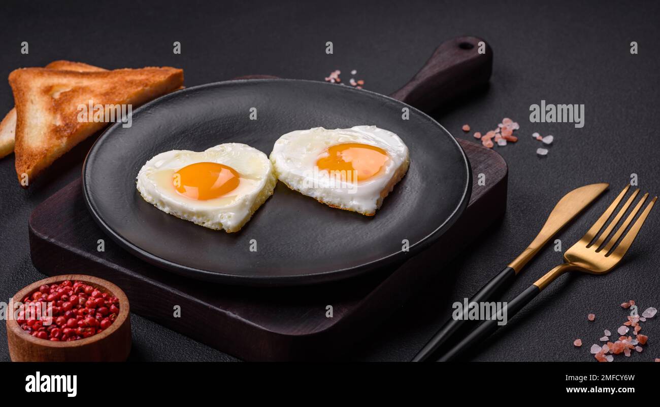 Two heart-shaped fried eggs on a black ceramic plate on a dark concrete ...
