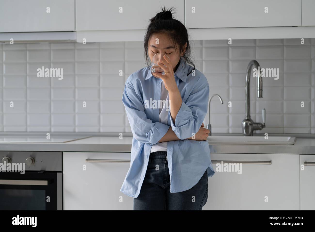 Stressed young Asian woman standing in kitchen drinking water calming ...