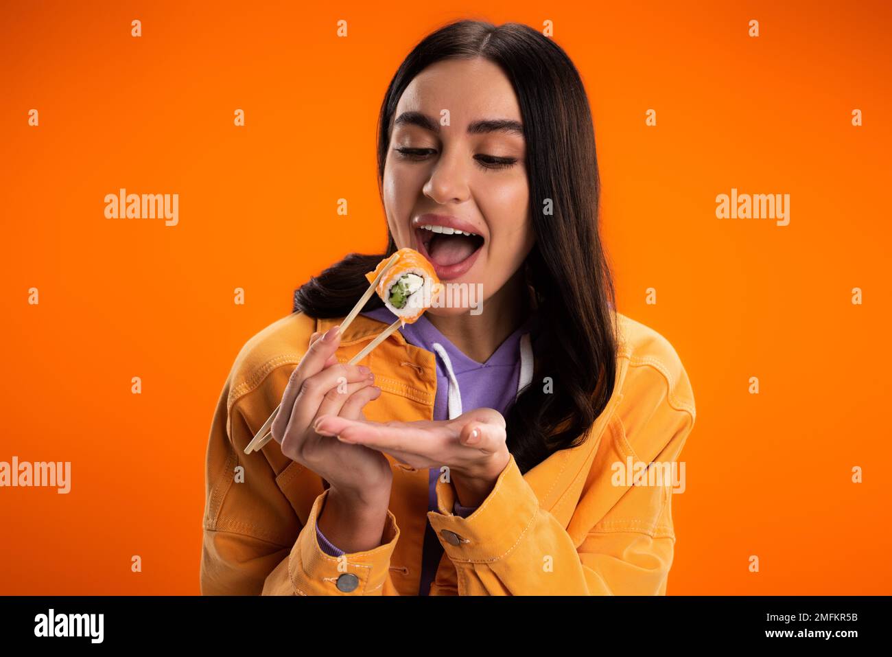 Brunette woman opening mouth and holding sushi isolated on orange,stock ...