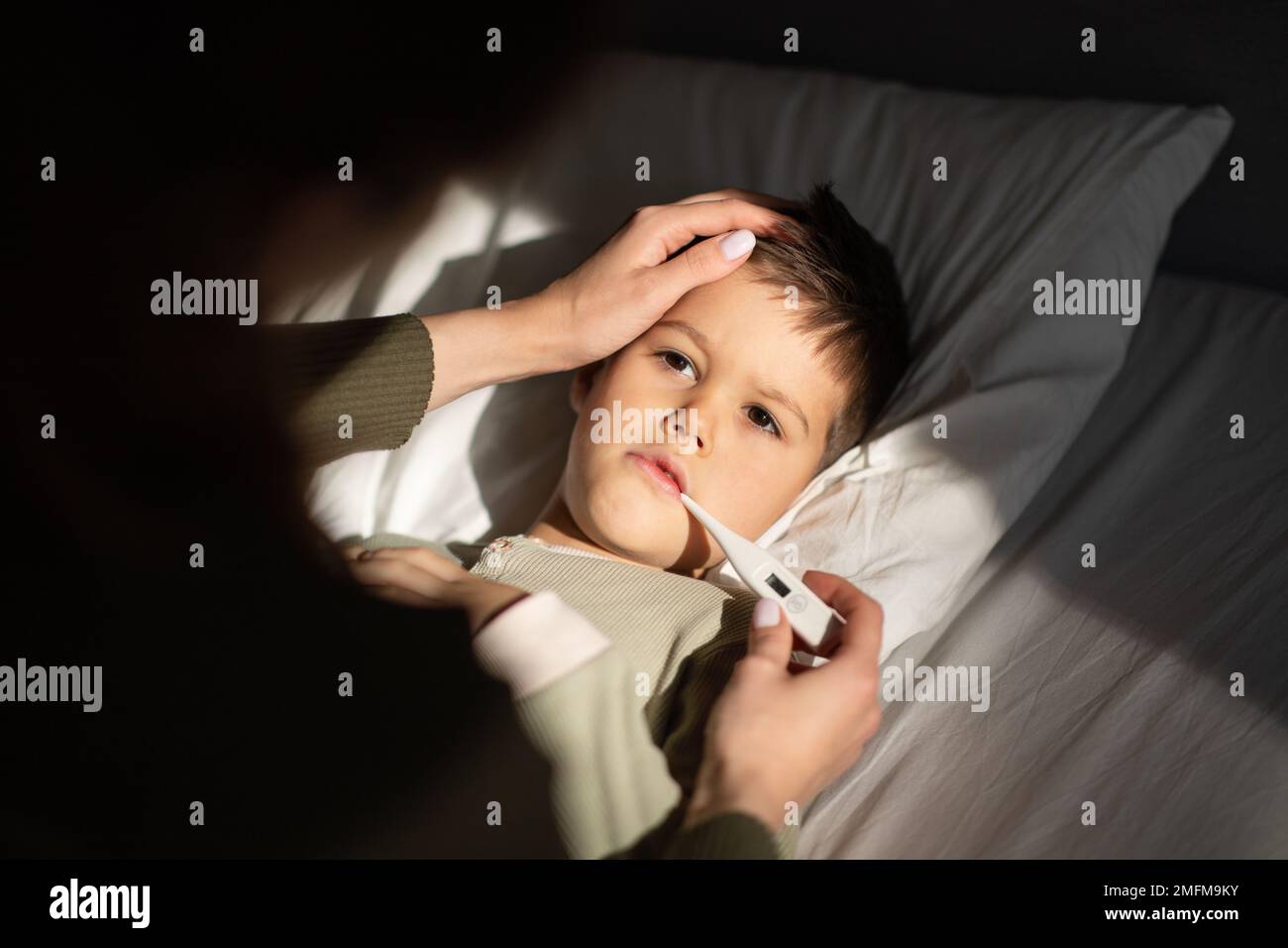 Sad little boy lies on bed, mom measures temperature with thermometer and touches forehead in ...