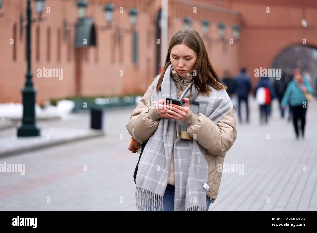Girl wearing down jacket and scarf standing with smartphone on a street ...