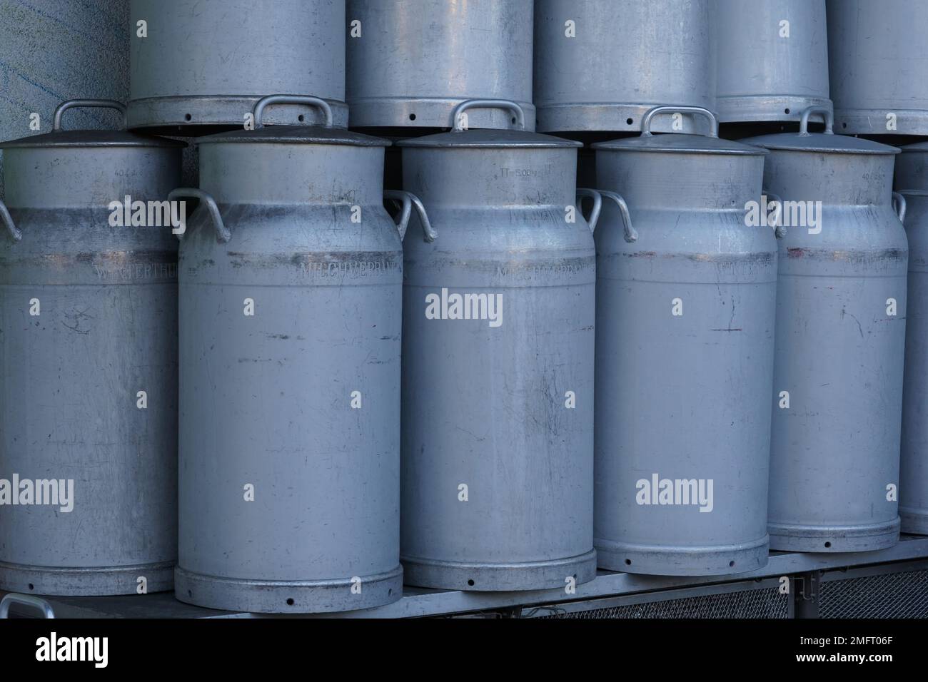 Milk churns made of metal stacked in rows. They have two handles and a ...
