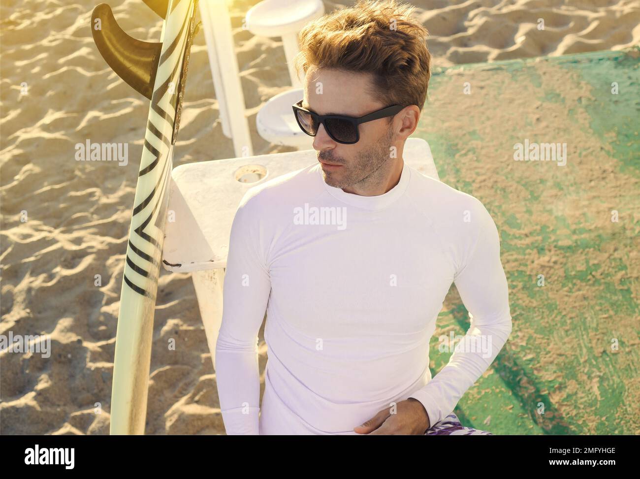Young handsome smiling man is sitting on a beach next to his surfboard ...