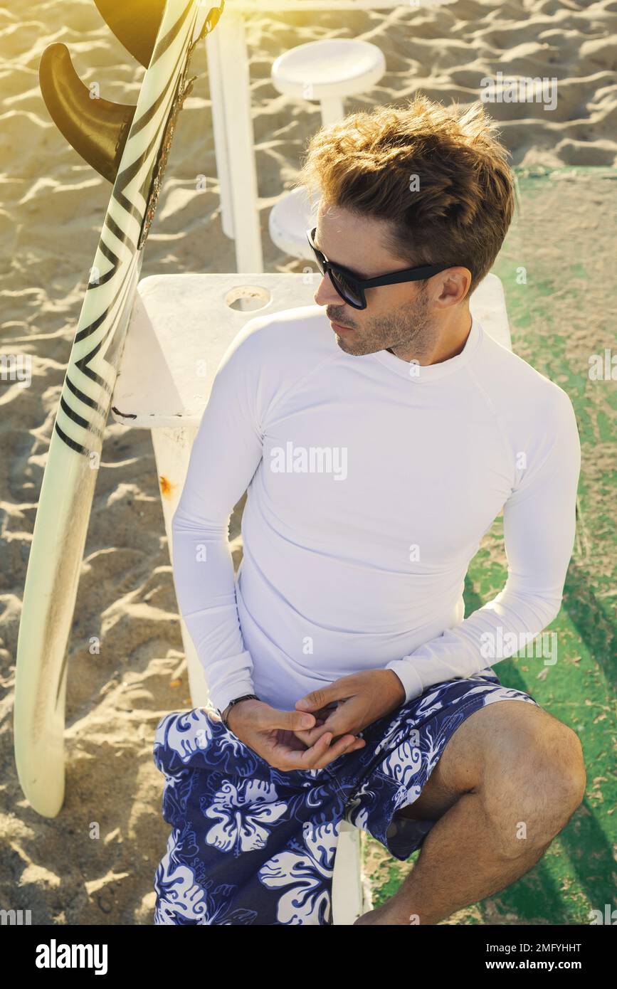 Young handsome smiling man is sitting on a beach next to his surfboard ...