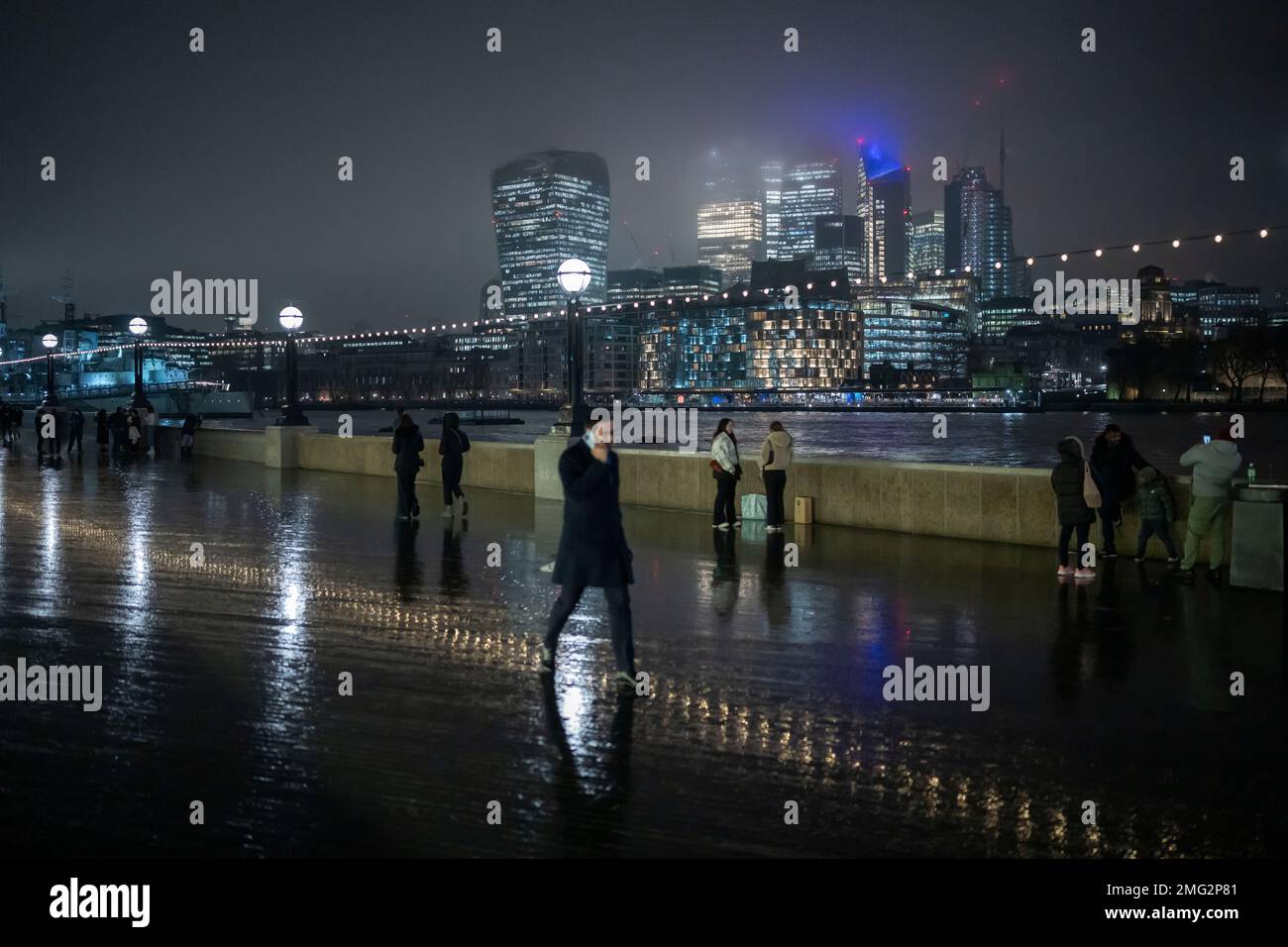 People walk along the bank of the River Thames with the skyline of the ...