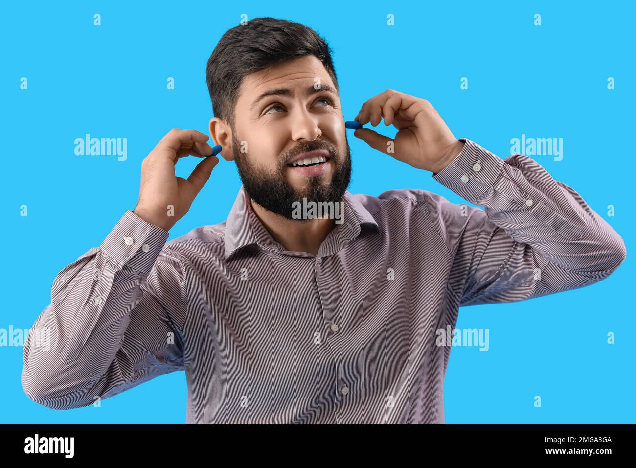 Young man with ear plugs suffering from loud noise on blue background ...