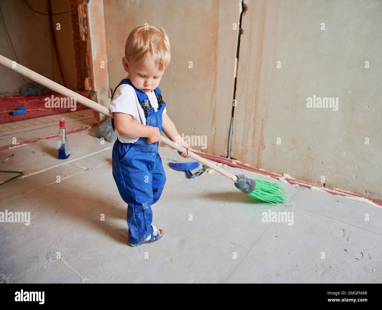 Full length of child in work overalls sweeping floor with broomstick at ...
