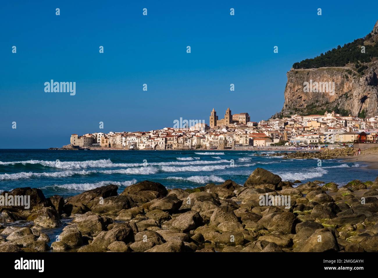 View across some rocks and the sea to the medieval town of Cefalu ...