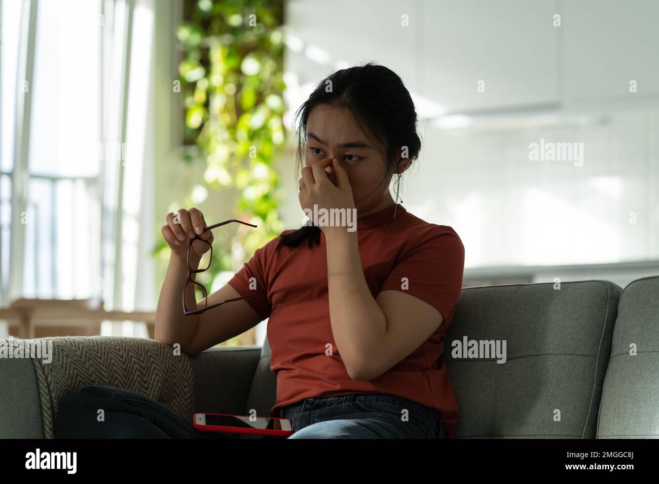 Tired Asian woman feeling sleepy while using smartphone at home, taking off glasses rubbing eyes ...