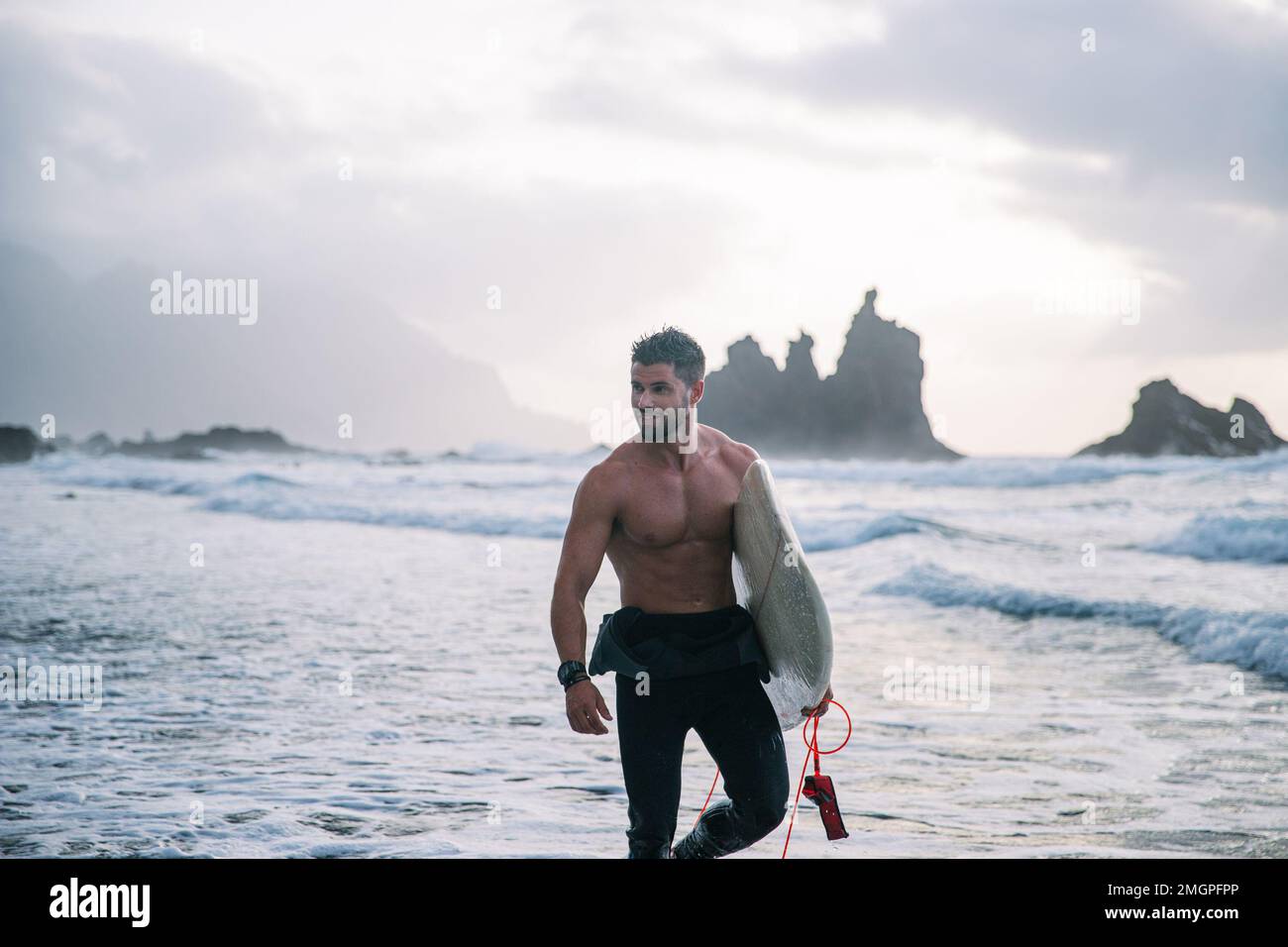 Surfer with surfboard walks on the beach, attractive guy looks around ...