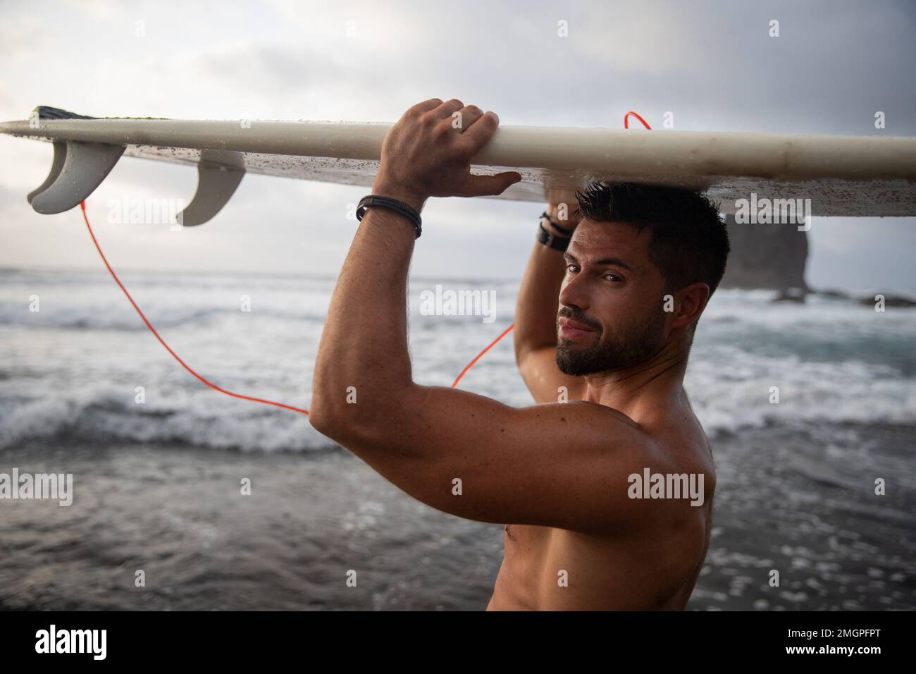 Attractive surfer rests surfboard on head and looks at the camera ...