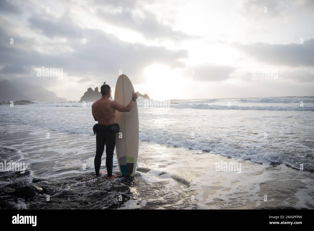 Surfer looks at the ocean while holding his surfboard at the beach ...