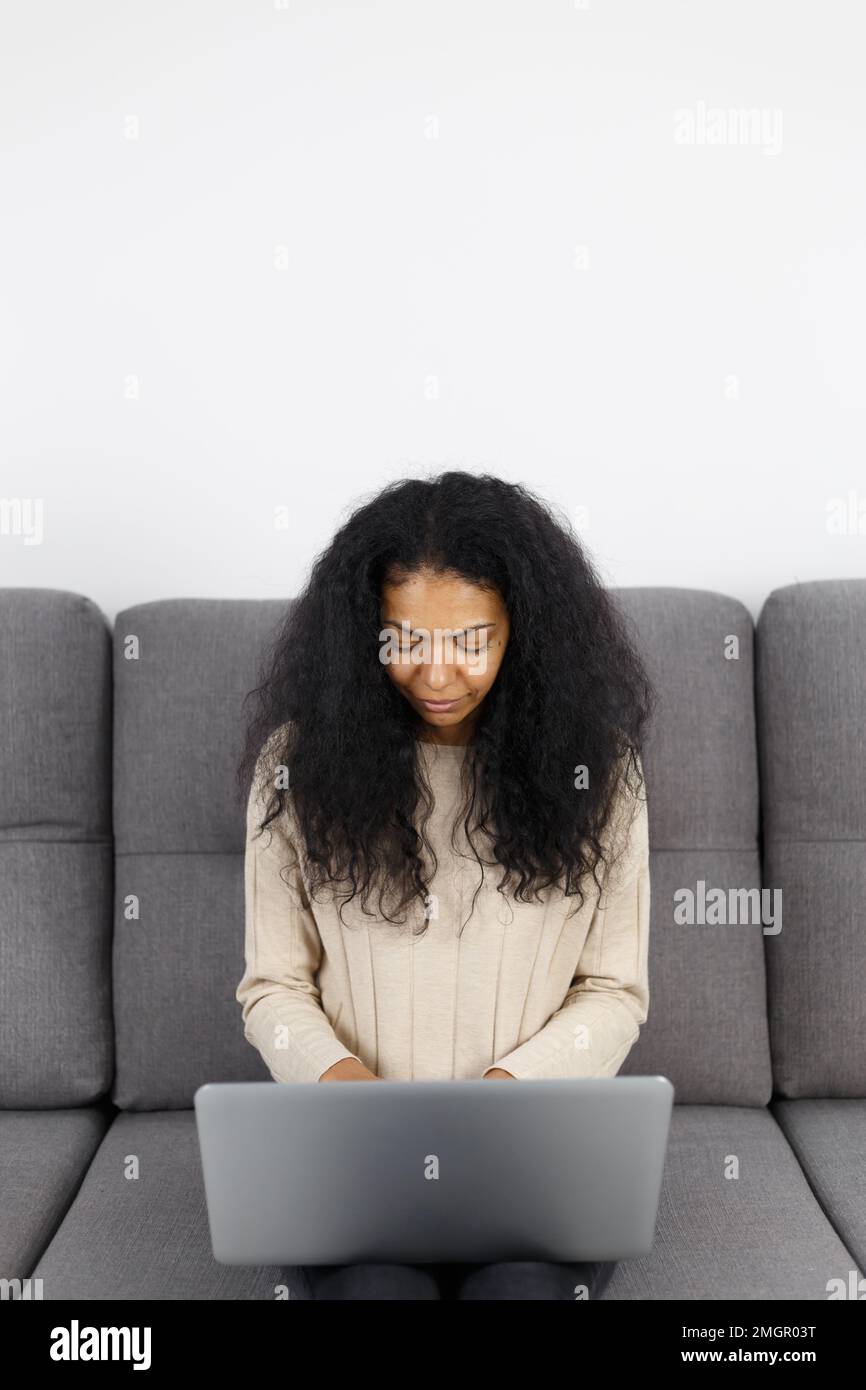BIPOC woman works on laptop on couch at home. POC female person doing ...