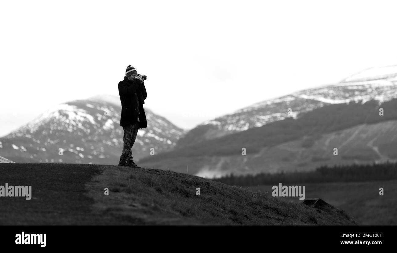 A grayscale shot of a Caucasian man taking pictures in a mountainous ...