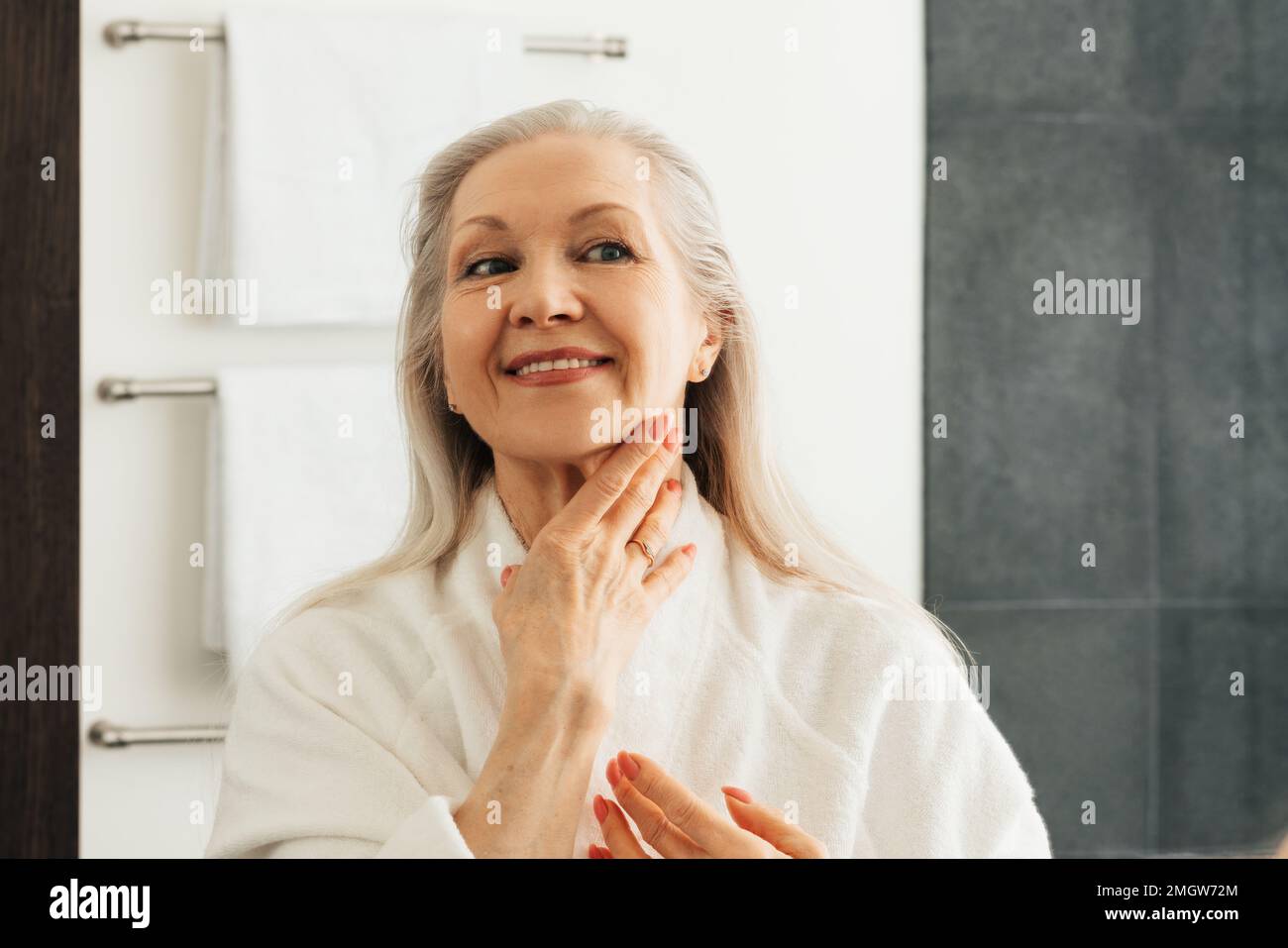 Smiling senior woman touching skin on her neck in front of a mirror ...
