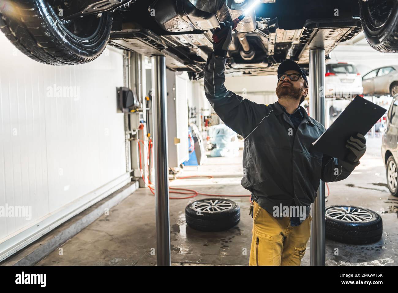 Mechanic with a clipboard inspecting the car chassis of a lifted car using a torch. High-quality ...
