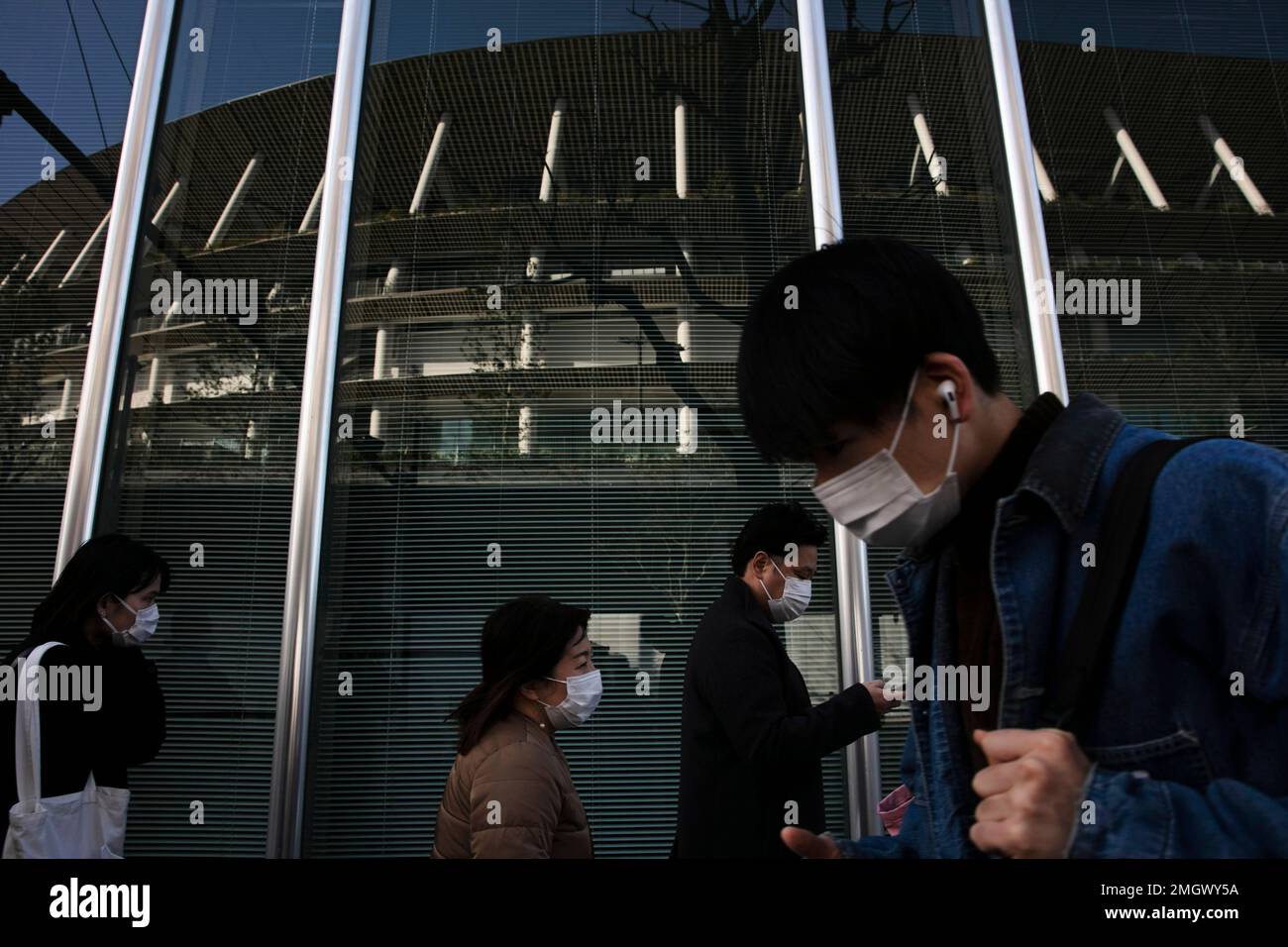 People with masks walk past a building reflecting the New National ...